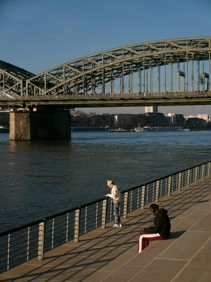 Two people sitting by Rhine River looking at Hohenzollern Bridge in Cologne