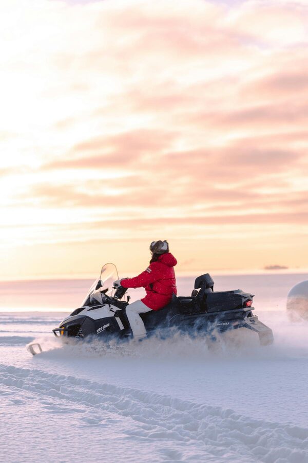 Person riding a snowmobile across a snowy field during a winter sunset